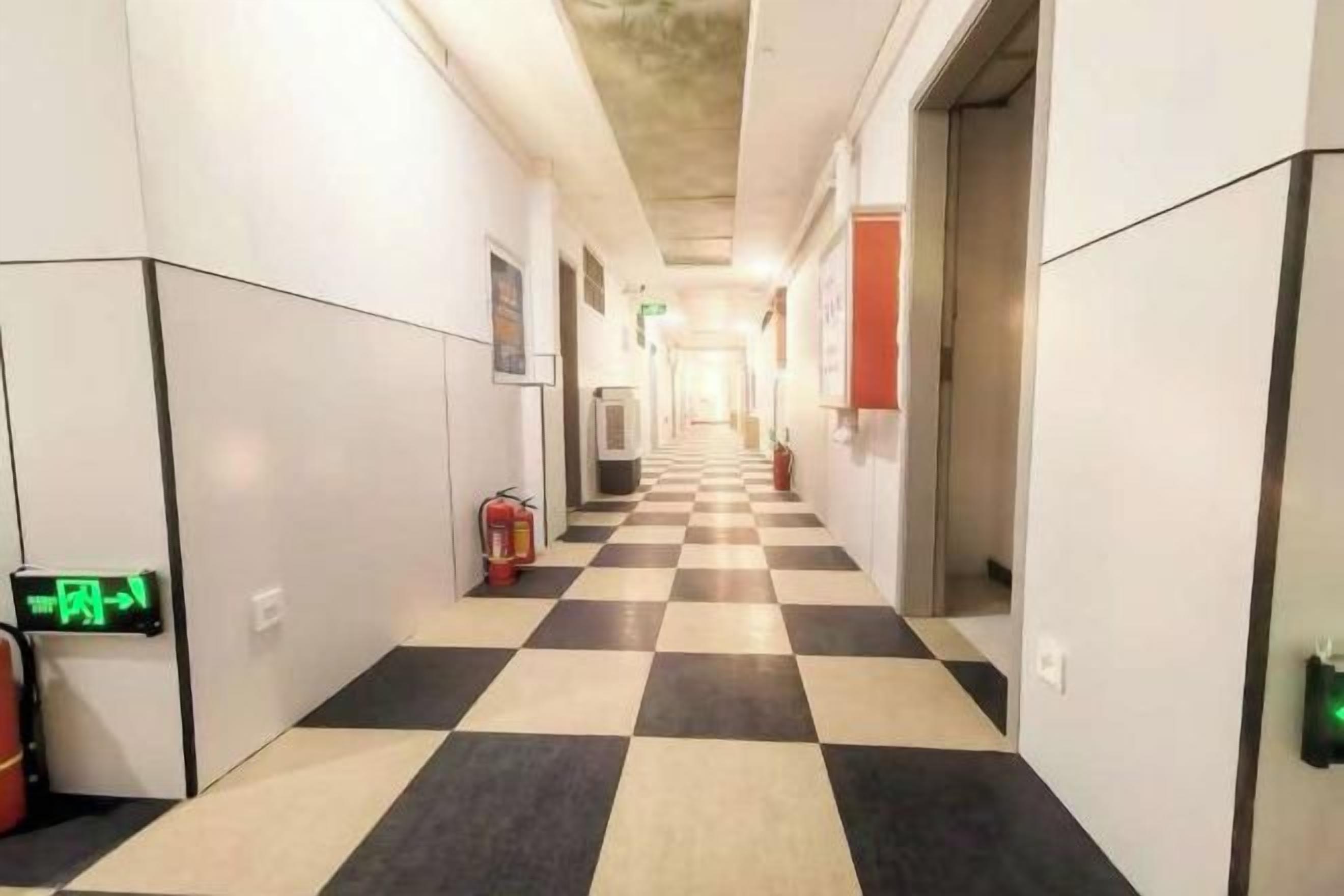 Corredor de hotel com piso xadrez, extintor de incêndio e placa de saída verde.