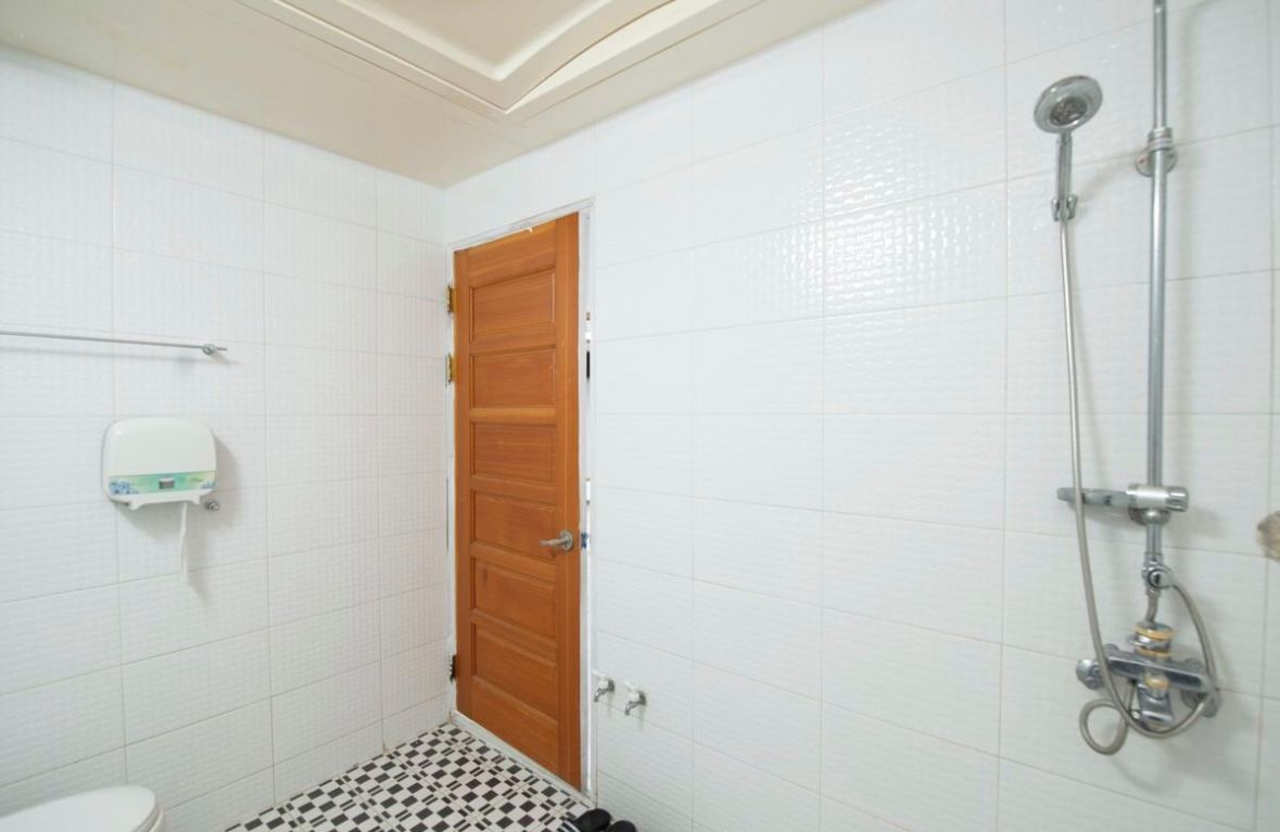 Casa de banho com paredes revestidas a azulejo branco, porta de madeira, sanita branca e chuveiro com acessório de duche manual.