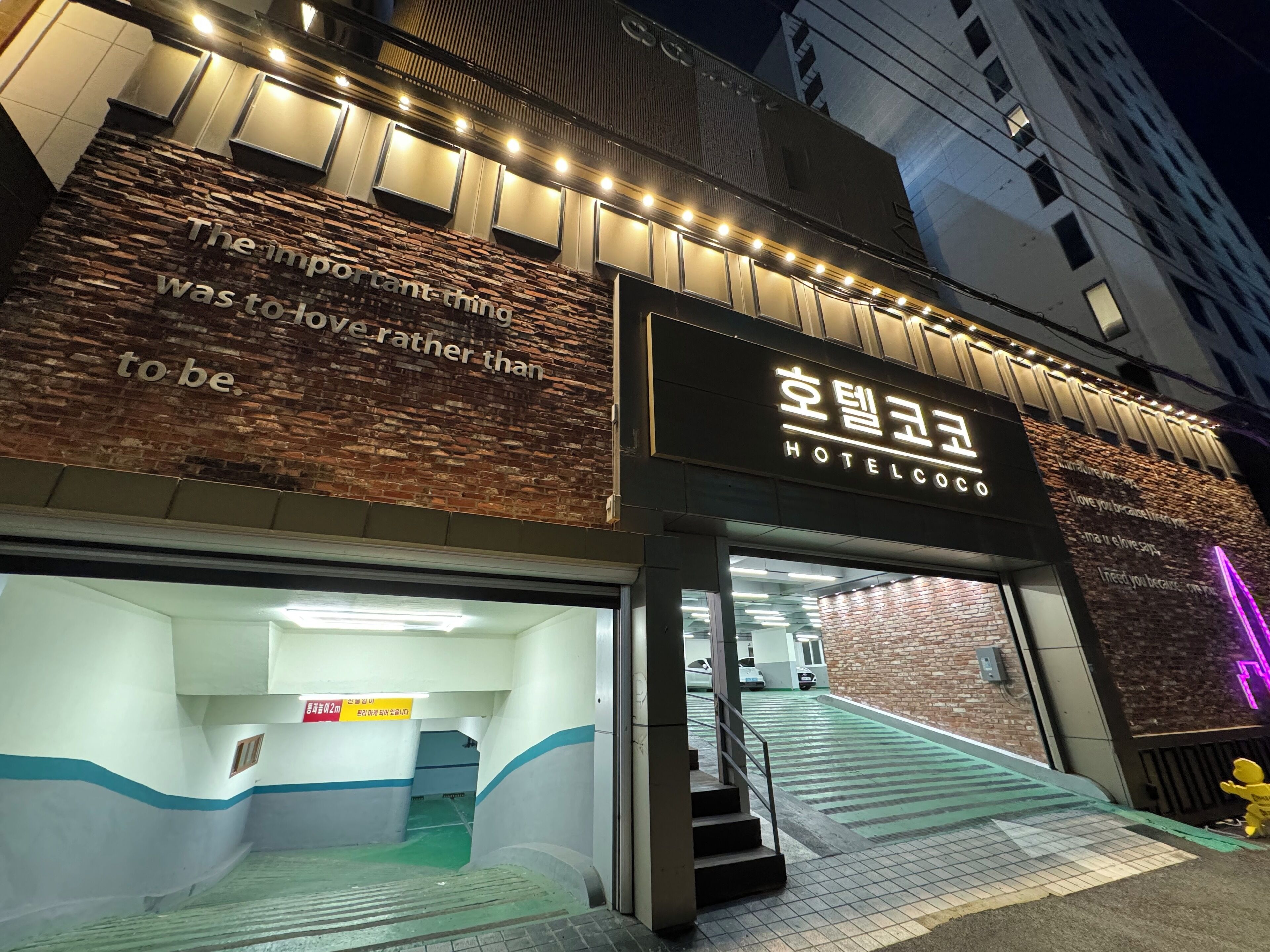 Entrada de hotel com fachada de tijolos, sinalização iluminada e escadaria que leva à entrada.