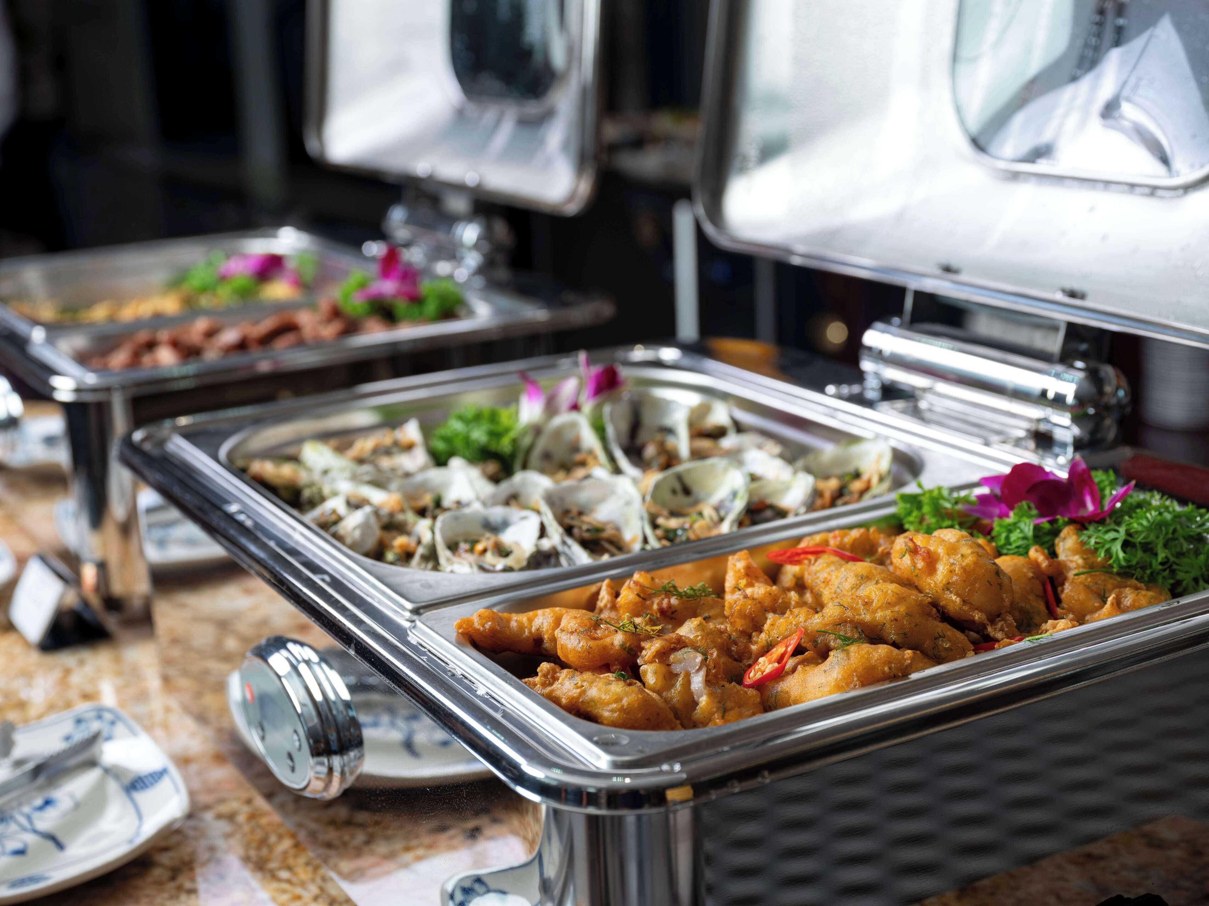 Serviço de buffet com diversos pratos, incluindo frutos do mar e frituras, dispostos em bandejas de metal.