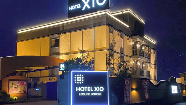 Um hotel moderno com uma placa grande 'HOTEL Xio', uma entrada bem iluminada e uma área ajardinada na frente.
