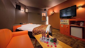 Quarto de hotel com cama, móveis laranja, uma mesa com garrafa e copos, televisão e minibar.