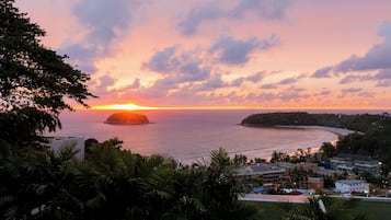 Vista de um pôr do sol costeiro com praia, palmeiras e edifícios em primeiro plano.