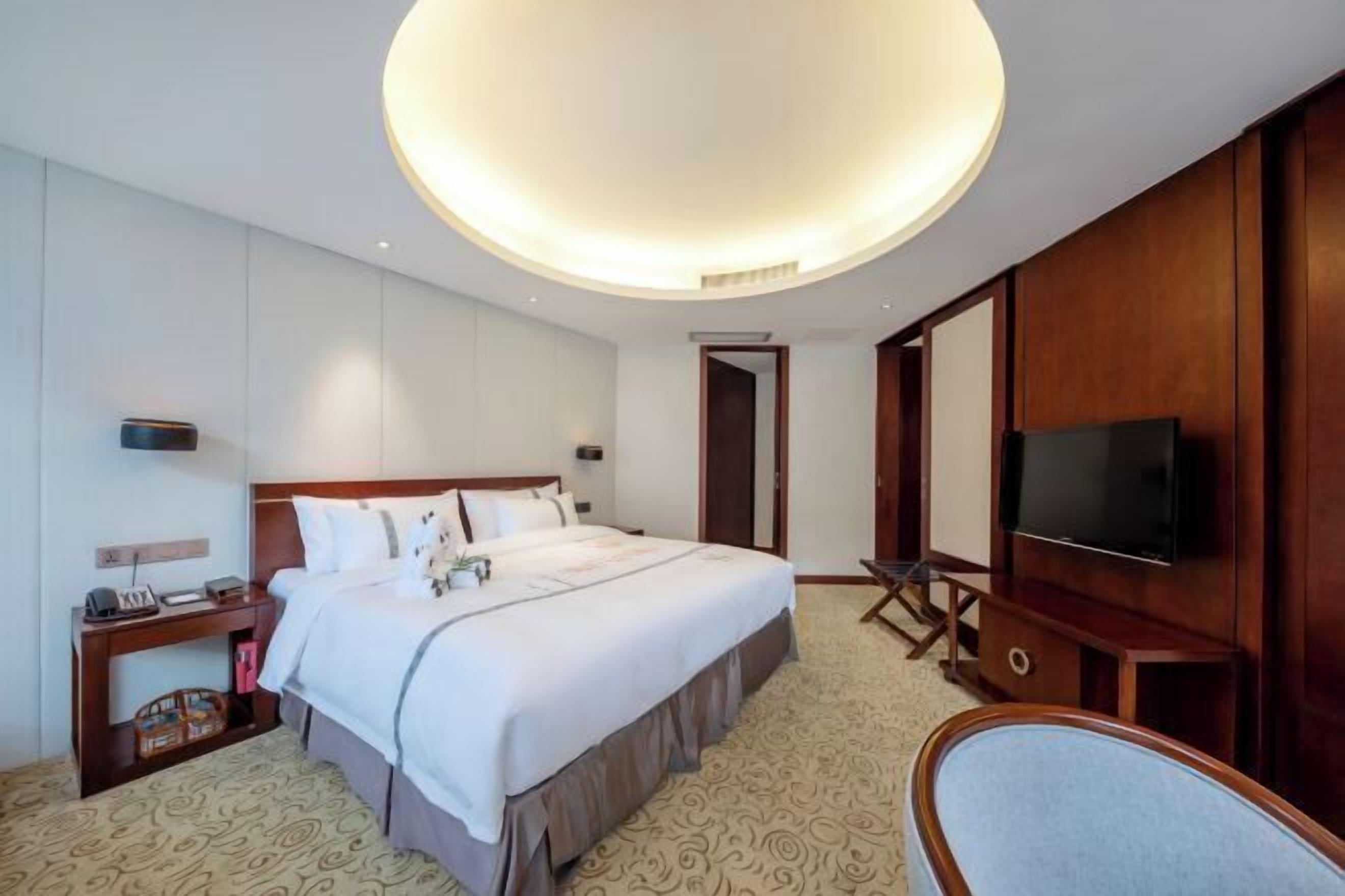 Quarto de hotel com uma cama grande, televisão, escrivaninha e luminária circular no teto.