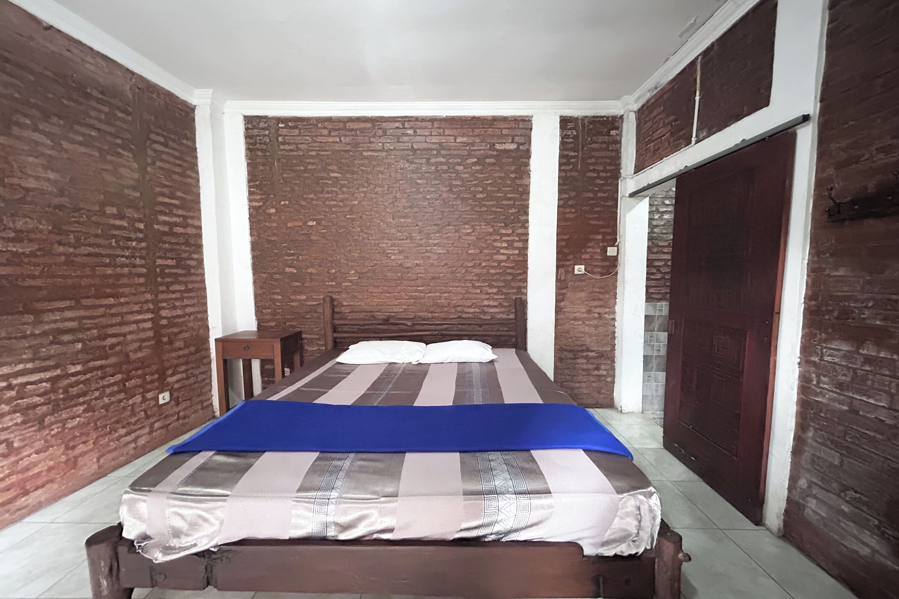 Quarto com cama, uma mesinha e uma cadeira. As paredes são de tijolo e há uma porta de madeira.