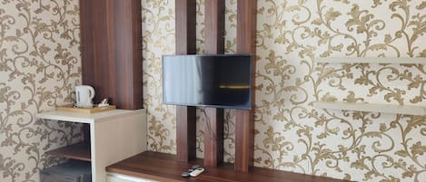 Quarto com televisão presa a um suporte de madeira, um frigorífico pequeno e uma prateleira com uma chaleira.