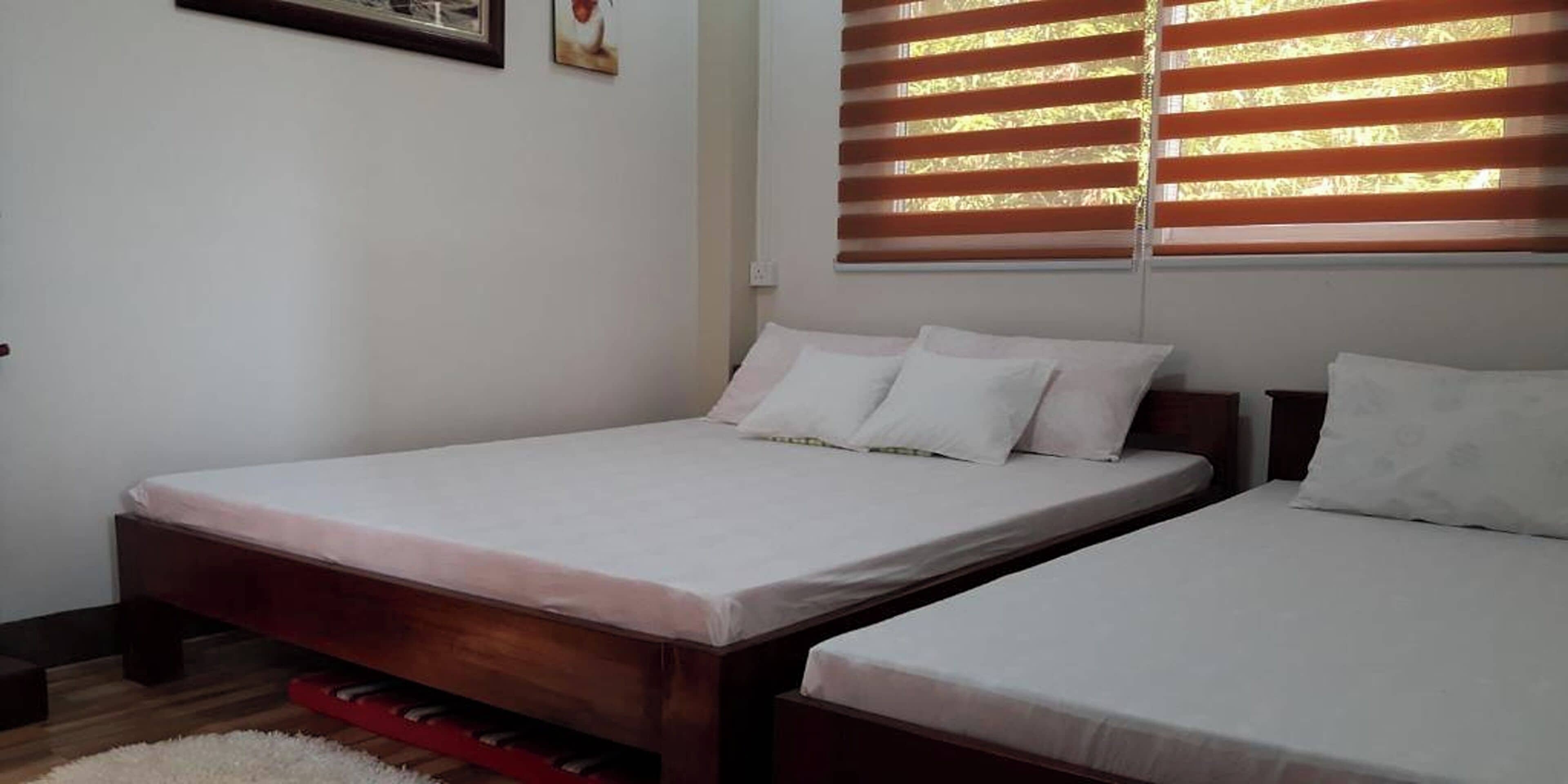 Quarto com duas camas individuais, cabeceiras de madeira, roupa de cama branca e uma janela com persianas.