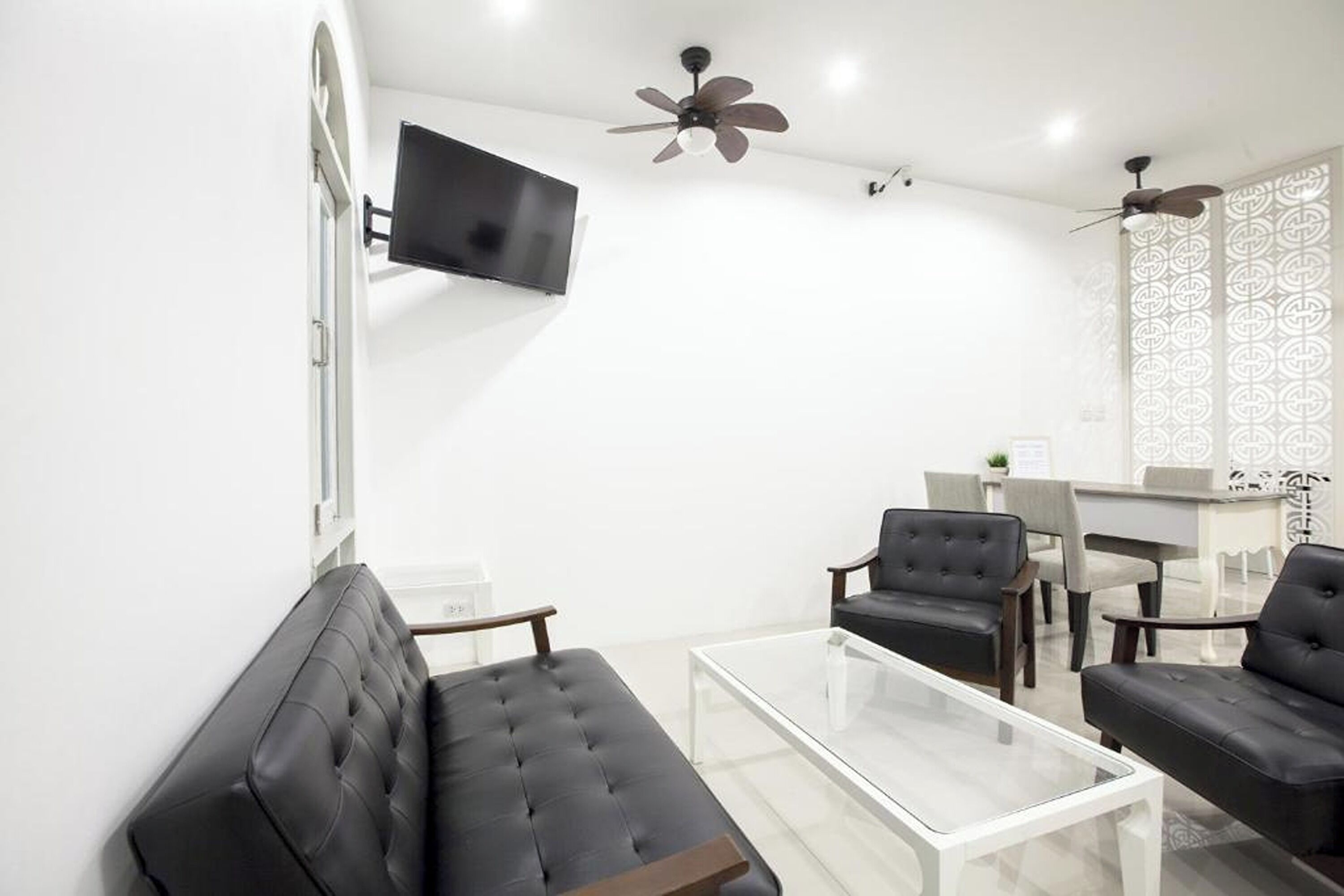 Uma sala de estar moderna com um sofá de couro preto, uma mesa de centro de vidro e duas poltronas. Há uma televisão de tela plana na parede e dois ventiladores de teto.