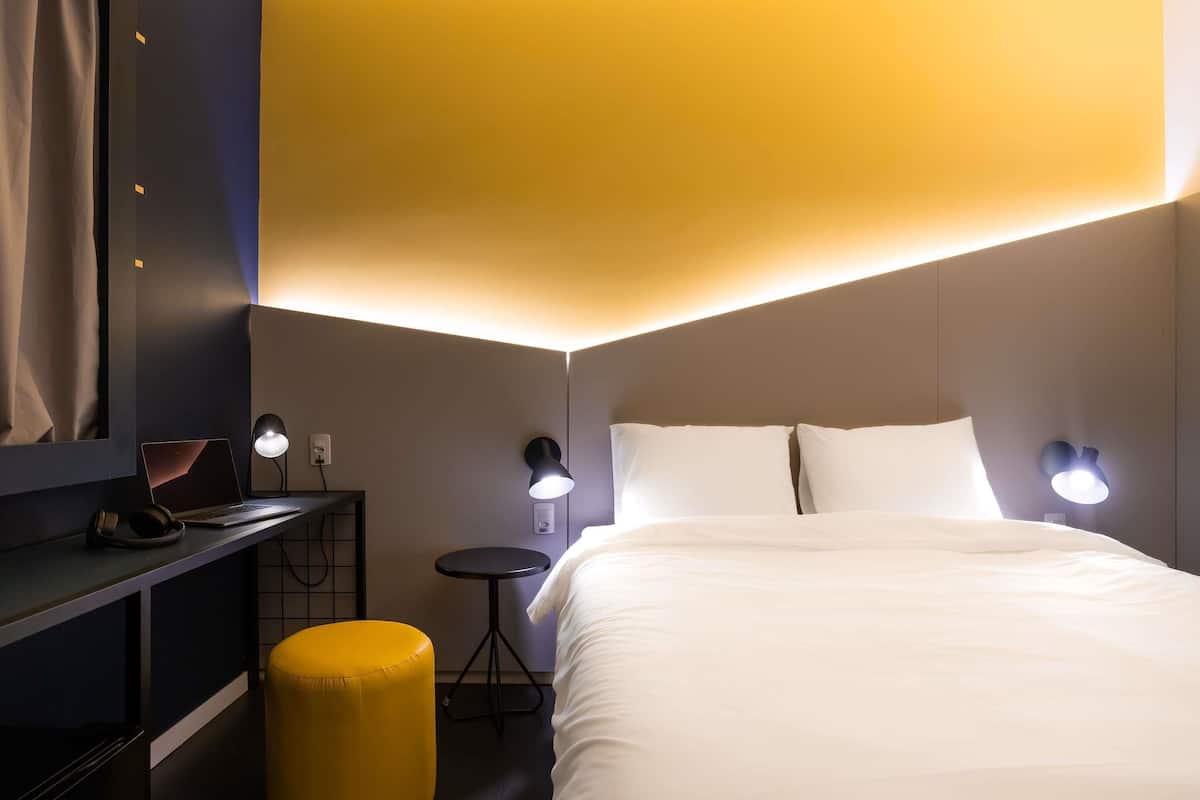 Quarto de hotel com cama, escrivaninha com laptop, um banquinho pequeno e duas luminárias fixadas na parede.