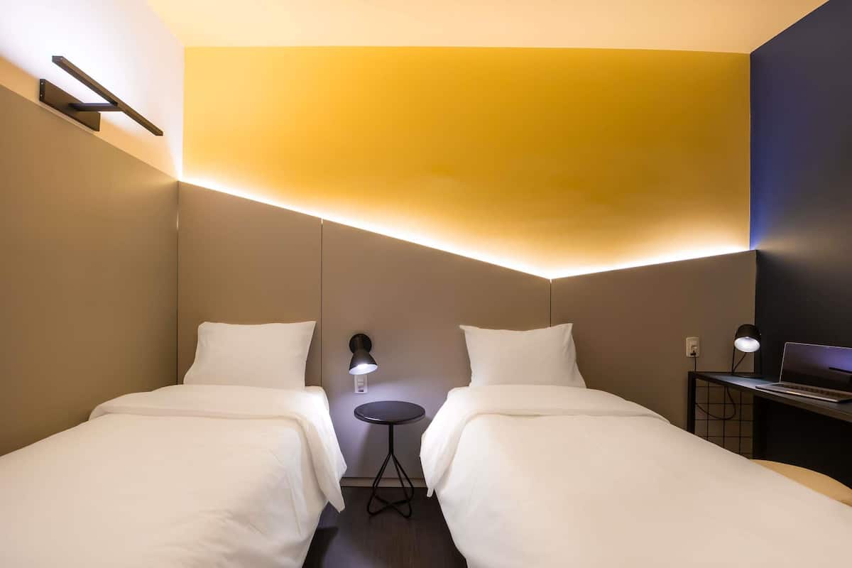 Quarto de hotel com duas camas, uma mesa redonda pequena, uma escrivaninha com um laptop e uma luminária fixada na parede.