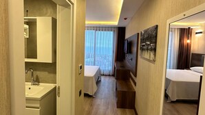 Corredor de hotel com um banheiro à esquerda, um quarto com cama e televisão à direita, e vista para a cidade pela janela.