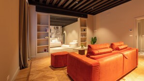 Uma sala de estar moderna com um sofá vermelho, um banquinho e uma mesa de centro. Há uma estante com diversos objetos e um quadro na parede.