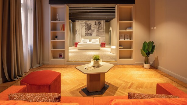 Quarto de hotel moderno com uma cama grande, área de estar com almofadas laranja, uma mesinha com uma planta e vista para o quarto através de uma porta.
