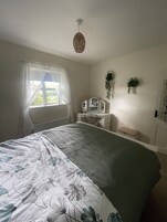 Quarto com cama, escrivaninha, cadeira, planta pendente e janela com cortinas.