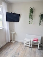 Quarto com televisão de tela plana na parede, um banco branco com almofada rosa e dois vasos suspensos com plantas verdes.