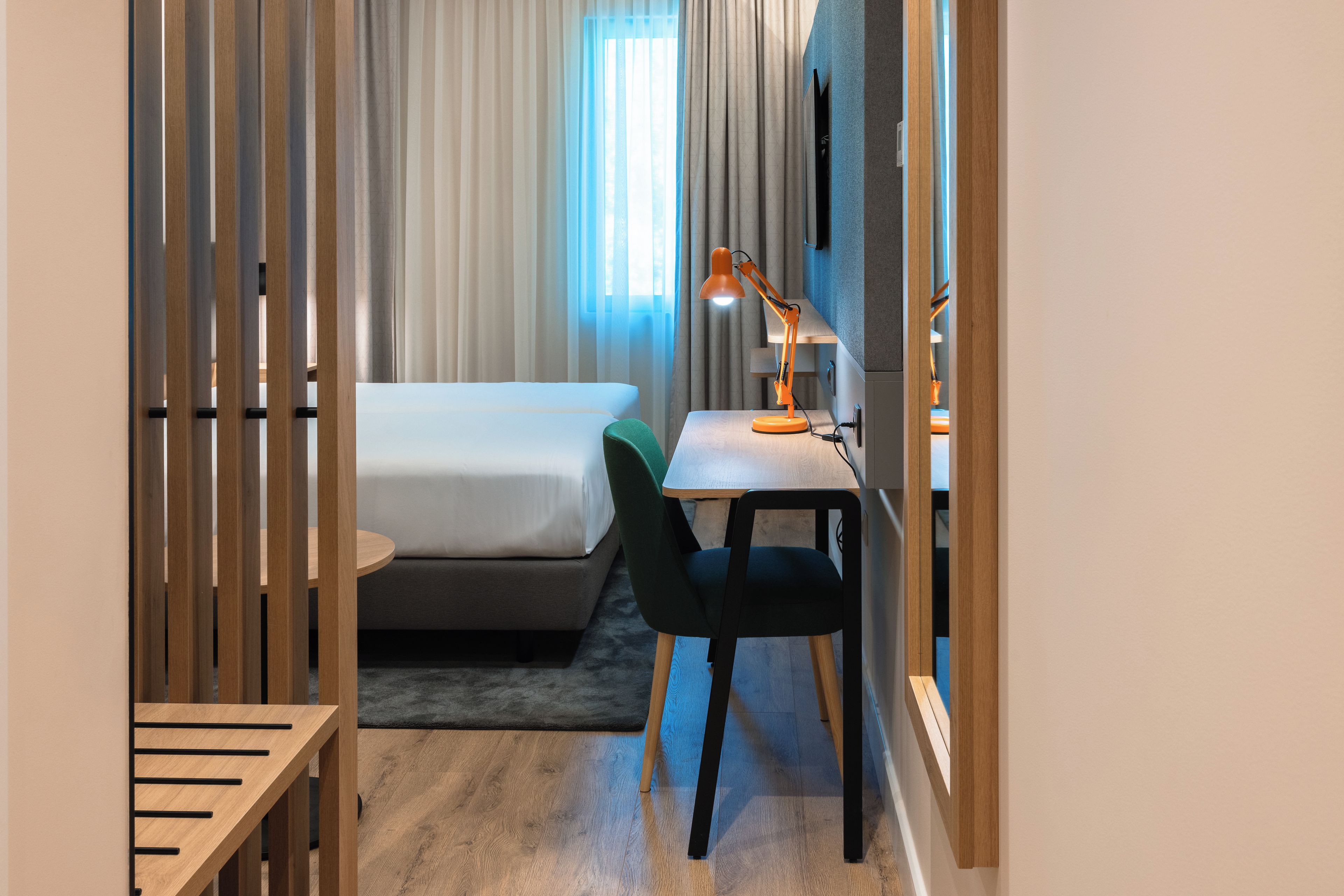 Quarto de hotel pequeno com uma cama, uma escrivaninha com luminária e uma cadeira.