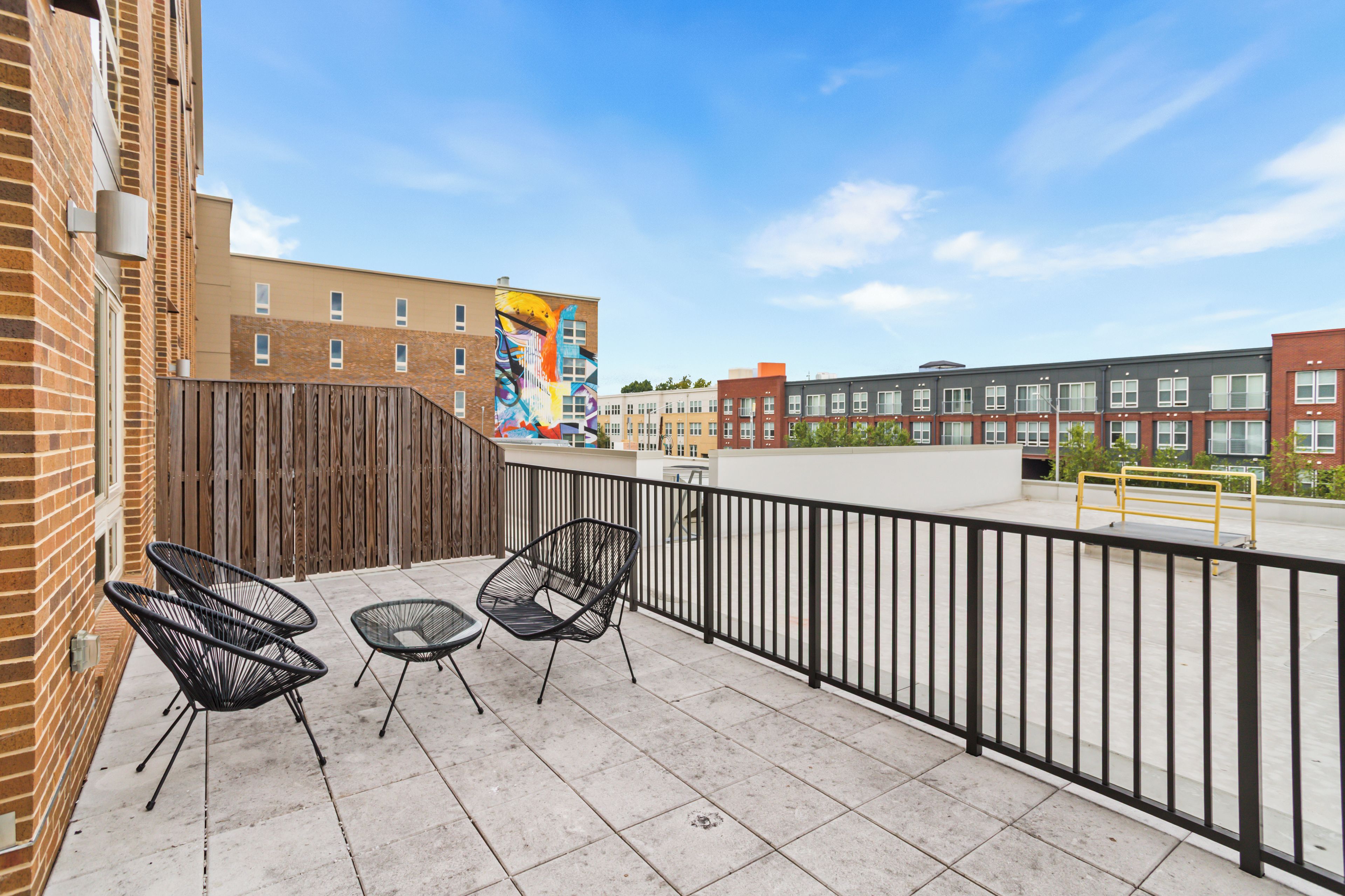 Um terraço no telhado com duas cadeiras e uma mesa, uma cerca de madeira e um mural em um edifício ao fundo.