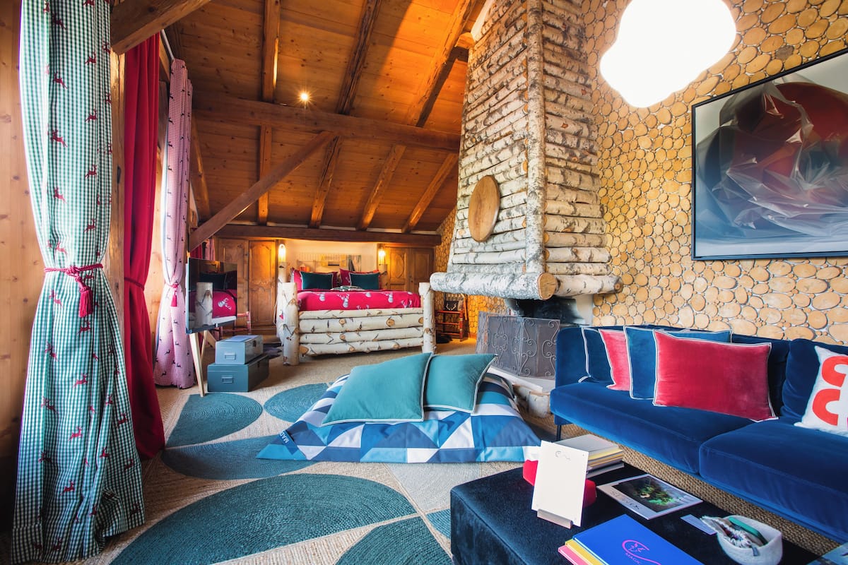 Um quarto aconchegante com lareira de pedra, um sofá azul com almofadas vermelhas e um tapete estampado.