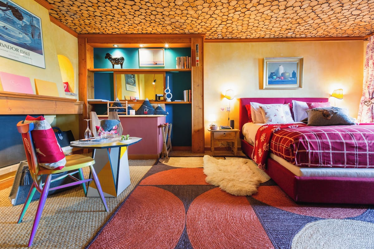 Quarto aconchegante com cama, escrivaninha e cadeira, destacando um design único no teto e um tapete colorido.