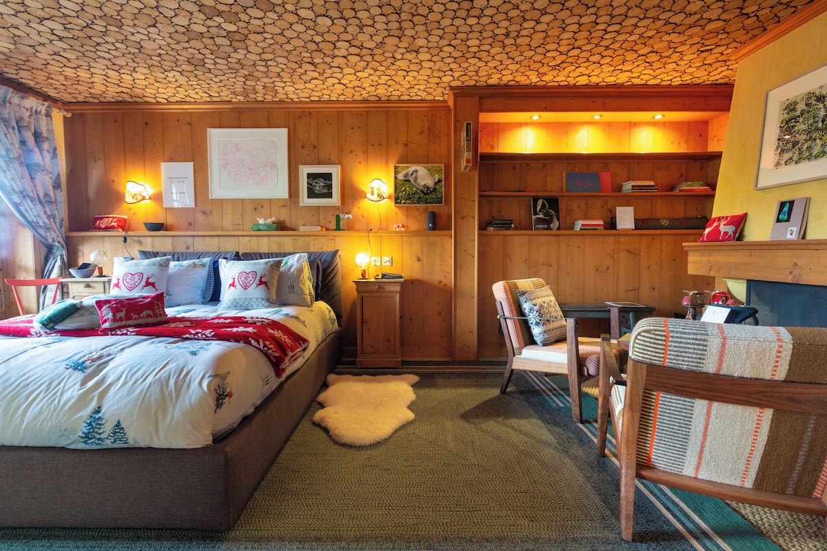 Quarto aconchegante com uma cama, uma poltrona, uma mesinha e uma estante com livros e objetos decorativos.