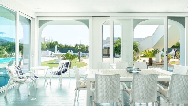 Um pátio amplo com móveis de vime branco, uma mesa de vidro e vista para uma piscina e vegetação.