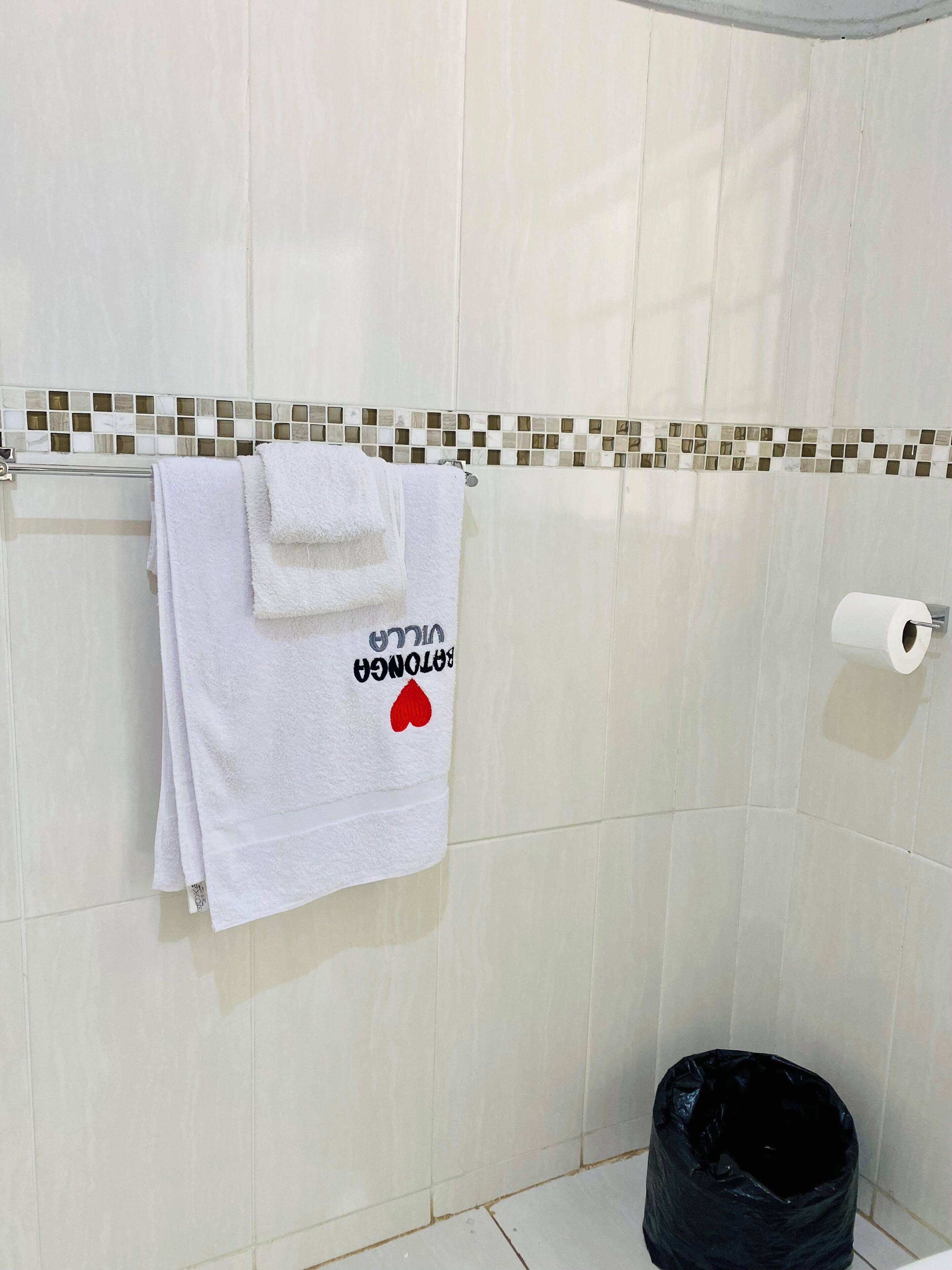 Casa de banho com paredes revestidas de azulejos brancos, um porta-toalhas com uma toalha branca, um rolo de papel higiénico e um caixote de lixo preto.