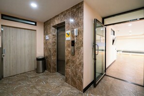 Um elevador com parede de mármore, uma lixeira e uma porta de vidro que dá para um corredor.