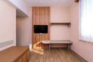 Quarto pequeno com um armário de madeira, uma televisão na parede, um banco e uma janela com cortinas.