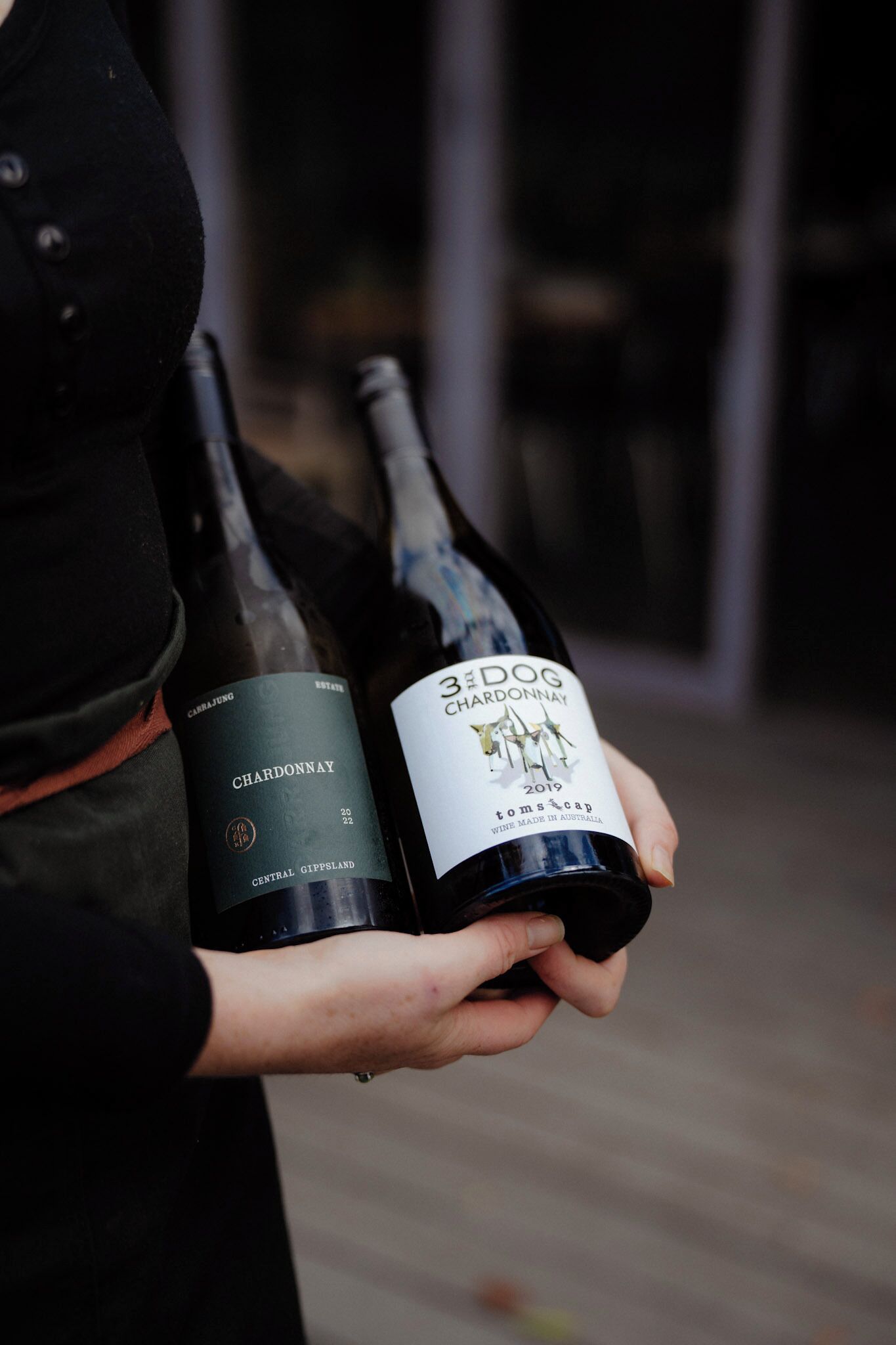 Uma pessoa segurando duas garrafas de vinho, uma delas com o rótulo 'Chardonnay' e a outra com o rótulo 'Bedog Chardonnay 2019'.