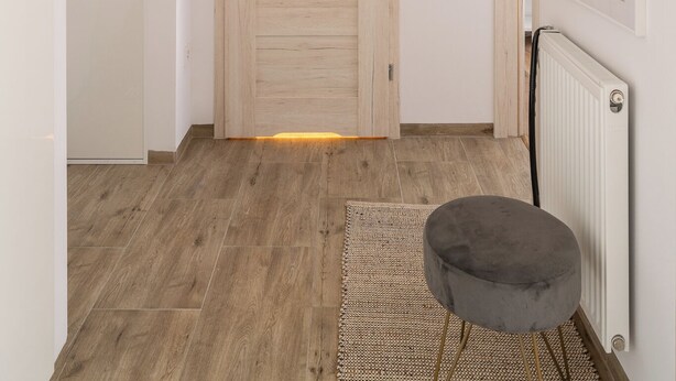 Um corredor com porta de madeira, radiador, um banquinho cinza e um quadro na parede.
