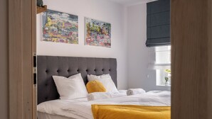 Quarto com cabeceira estofada cinza, roupa de cama branca, manta amarela mostarda e duas pinturas coloridas com paisagens urbanas na parede.