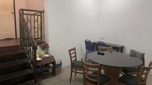 Uma área de refeições com uma mesa redonda, quatro cadeiras de madeira, uma escada com guarda-corpo metálico e uma pequena lixeira.
