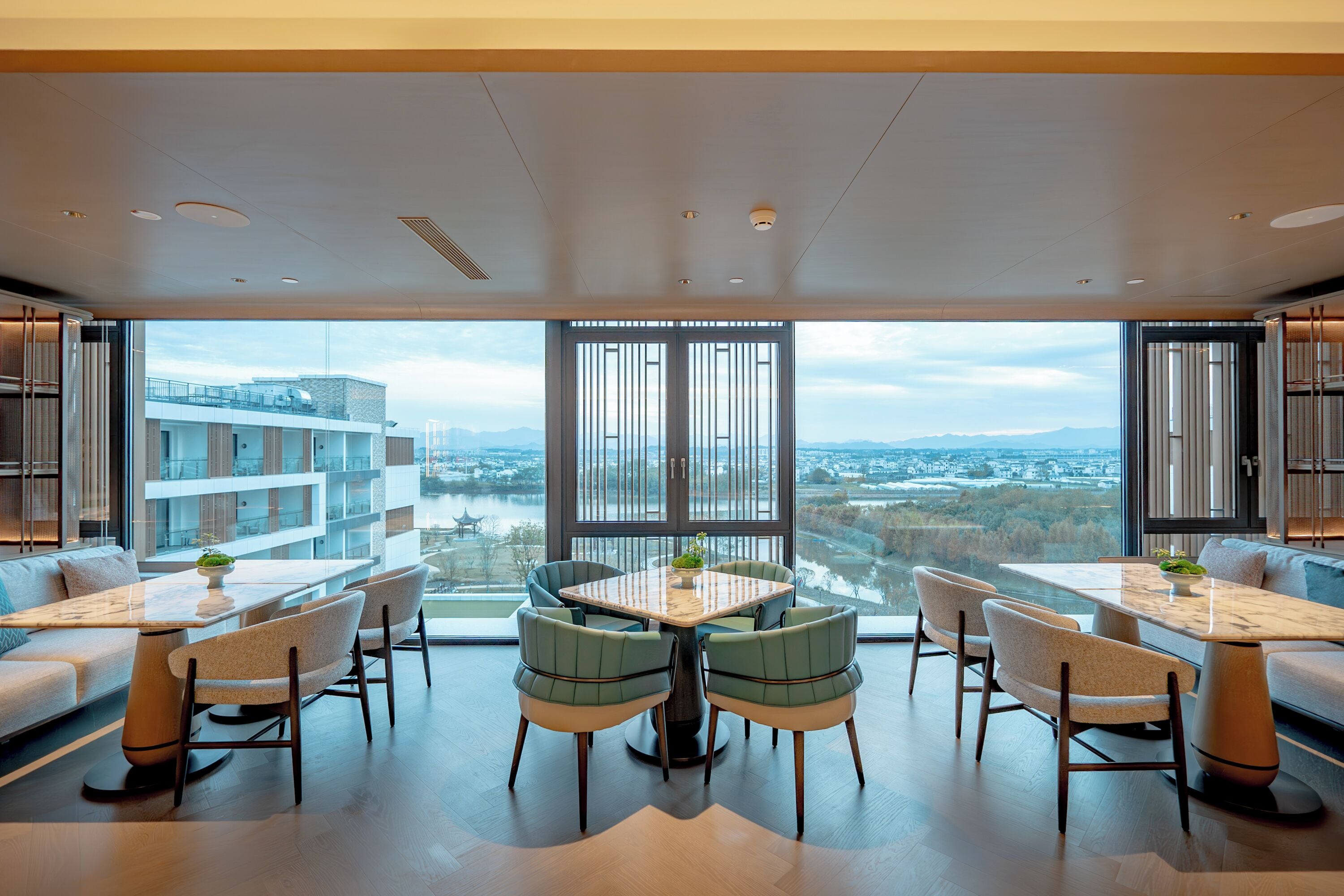 Quarto de hotel moderno com uma grande janela, área de refeições com mesa e cadeiras, e um sofá.