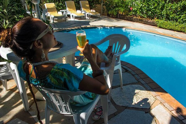 Uma pessoa sentada à beira da piscina com uma bebida, rodeada por espreguiçadeiras e vegetação.