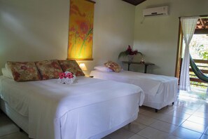 Quarto de hotel com duas camas, uma grande tapeçaria floral na parede, aparelho de ar condicionado, uma mesa pequena com um vaso de flores e uma rede lá fora.