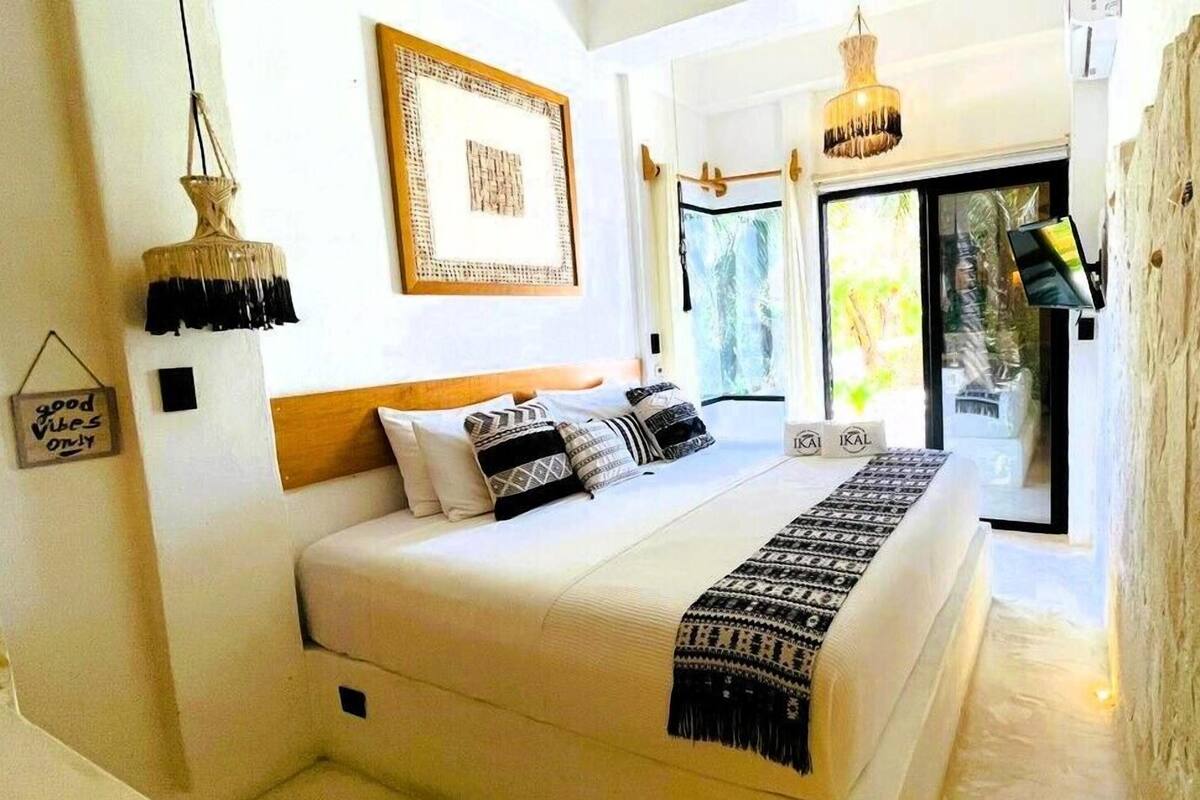 Quarto com uma cama grande, cabeceira de madeira, manta estampada e luminária pendente.