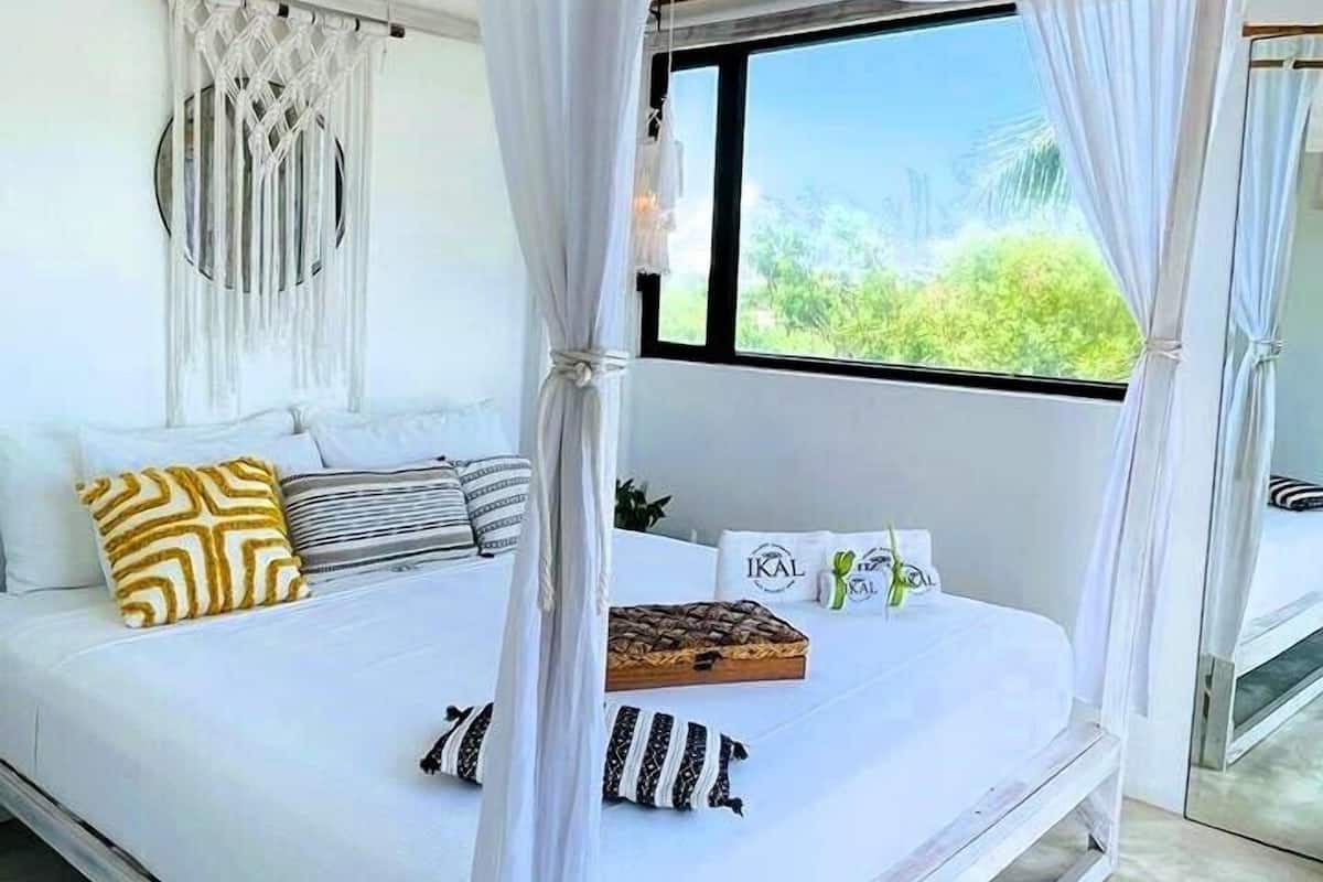 Uma cama de dossel com colchas e almofadas brancas, um ventilador de teto e vista para o verde lá fora.