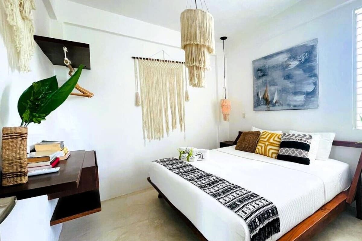 Quarto moderno com uma cama grande, uma mesinha de cabeceira, uma decoração de macramé pendurada e um quadro na parede.