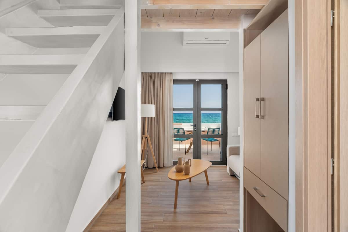 Quarto de hotel moderno com escada, piso de madeira, sofá, mesa com objetos decorativos e vista para o mar através de uma porta de vidro.