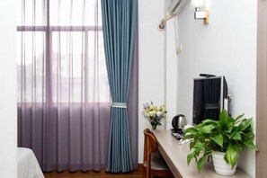 Quarto de hotel com cama, escrivaninha com computador, planta em vaso e janela com cortinas leves.