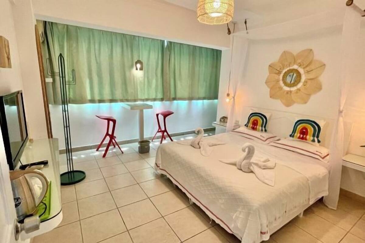 Quarto de hotel com uma cama, uma mesinha, dois taburetes vermelhos, um espelho e uma janela com cortinas verdes.