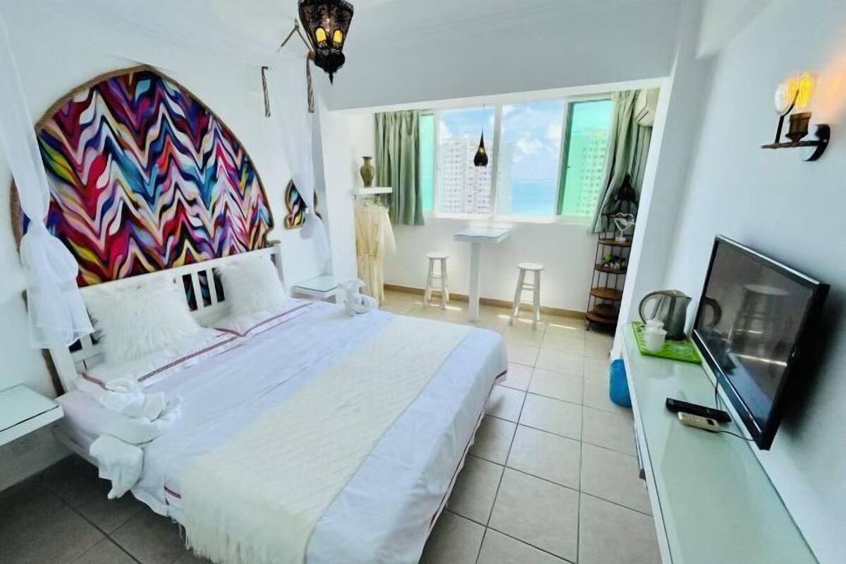 Quarto com cama, escrivaninha, cadeira, televisão e janela com vista.