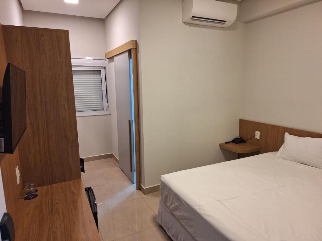 Quarto de hotel com cama, guarda-roupa de madeira, mesa de cabeceira, televisão e janela com persianas.