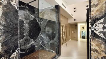 Interior moderno com área de chuveiro em vidro, paredes de mármore e um banco de madeira.