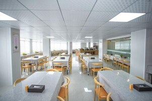Uma sala de jantar ampla com várias mesas e cadeiras, uma cozinha ao fundo e teto com azulejos.
