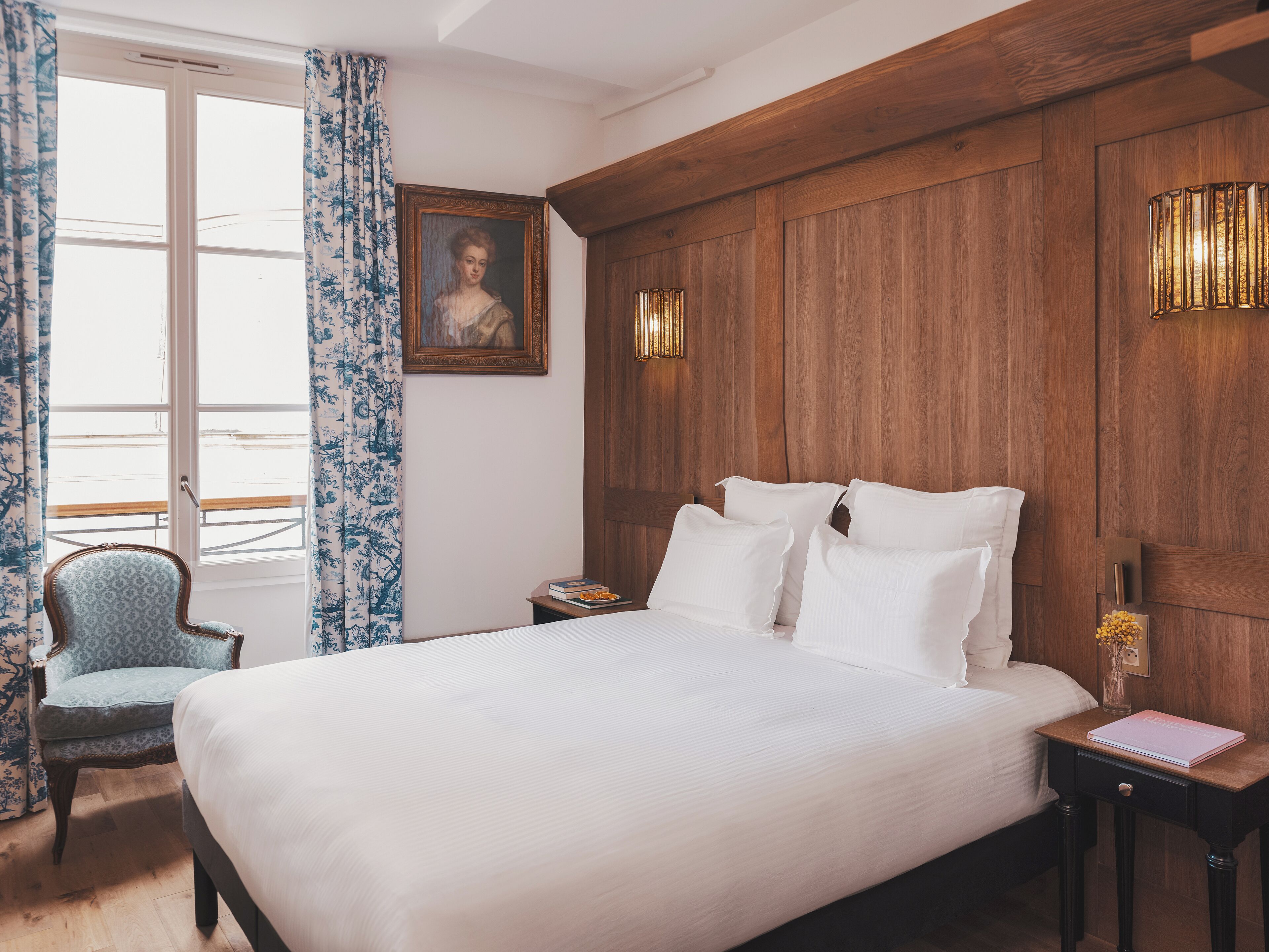 Quarto com uma cama grande, cabeceira de madeira, mesa de cabeceira com abajur, uma cadeira e uma janela com cortinas.