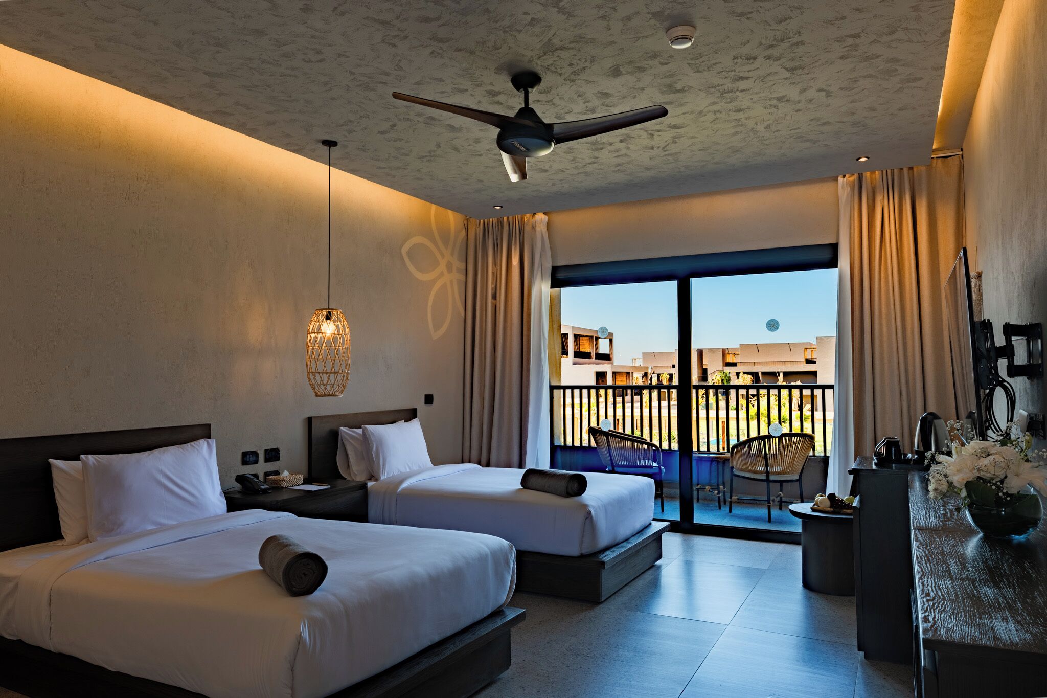 Quarto de hotel com duas camas, ventilador de teto, varanda com vista e escrivaninha com um vaso de flores.
