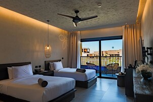 Quarto de hotel com duas camas, ventilador de teto, varanda com vista e escrivaninha com um vaso de flores.