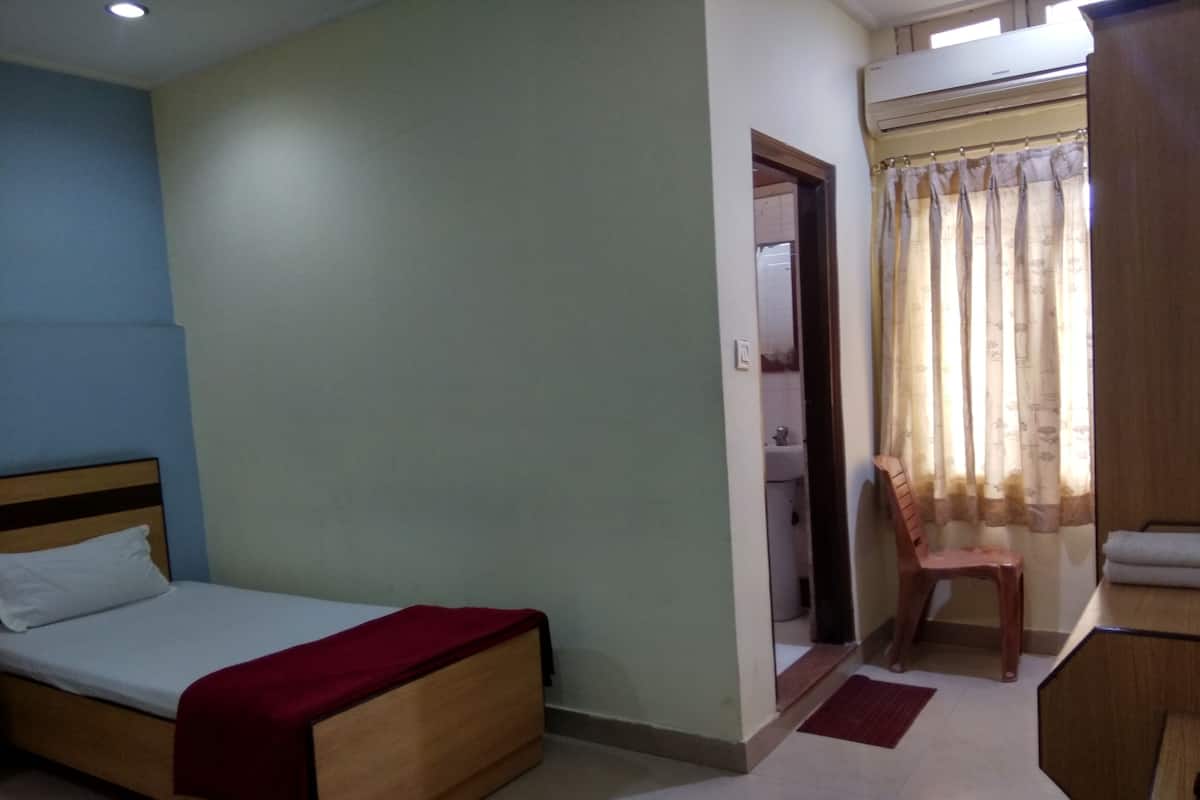 Um quarto com uma cama, cabeceira de madeira, manta vermelha, uma cadeira de madeira, uma pequena mesa com uma toalha e um banheiro visível através de uma porta aberta.