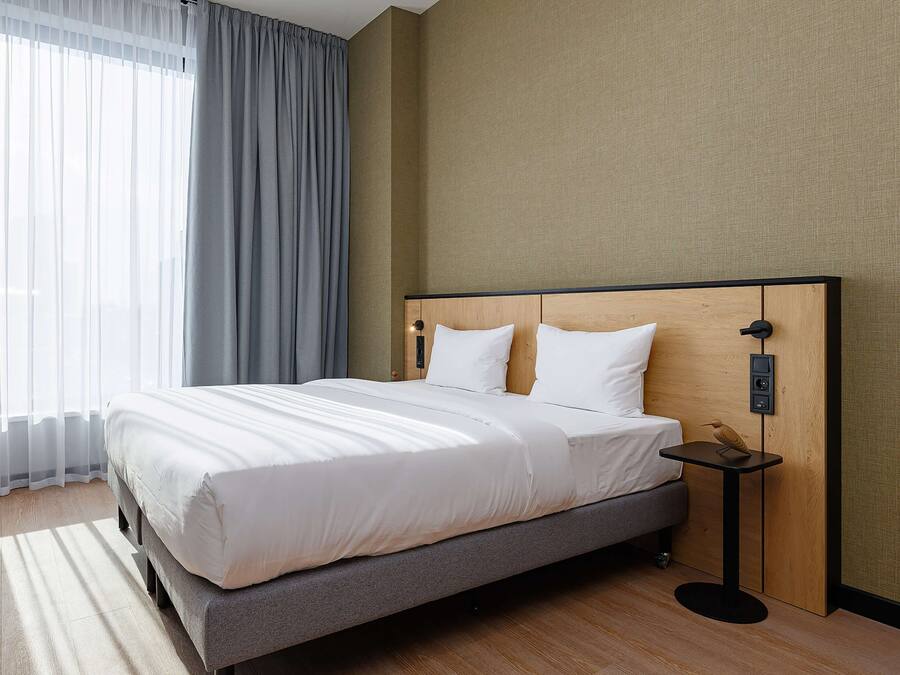 Quarto de hotel com uma cama grande, uma mesinha de cabeceira e vista para o exterior através de cortinas translúcidas.